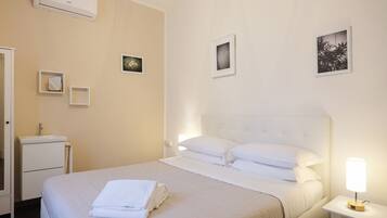Quarto de hotel com cama, mesa de cabeceira com abajur, criado-mudo e ar-condicionado fixado na parede, além de quadros na parede.