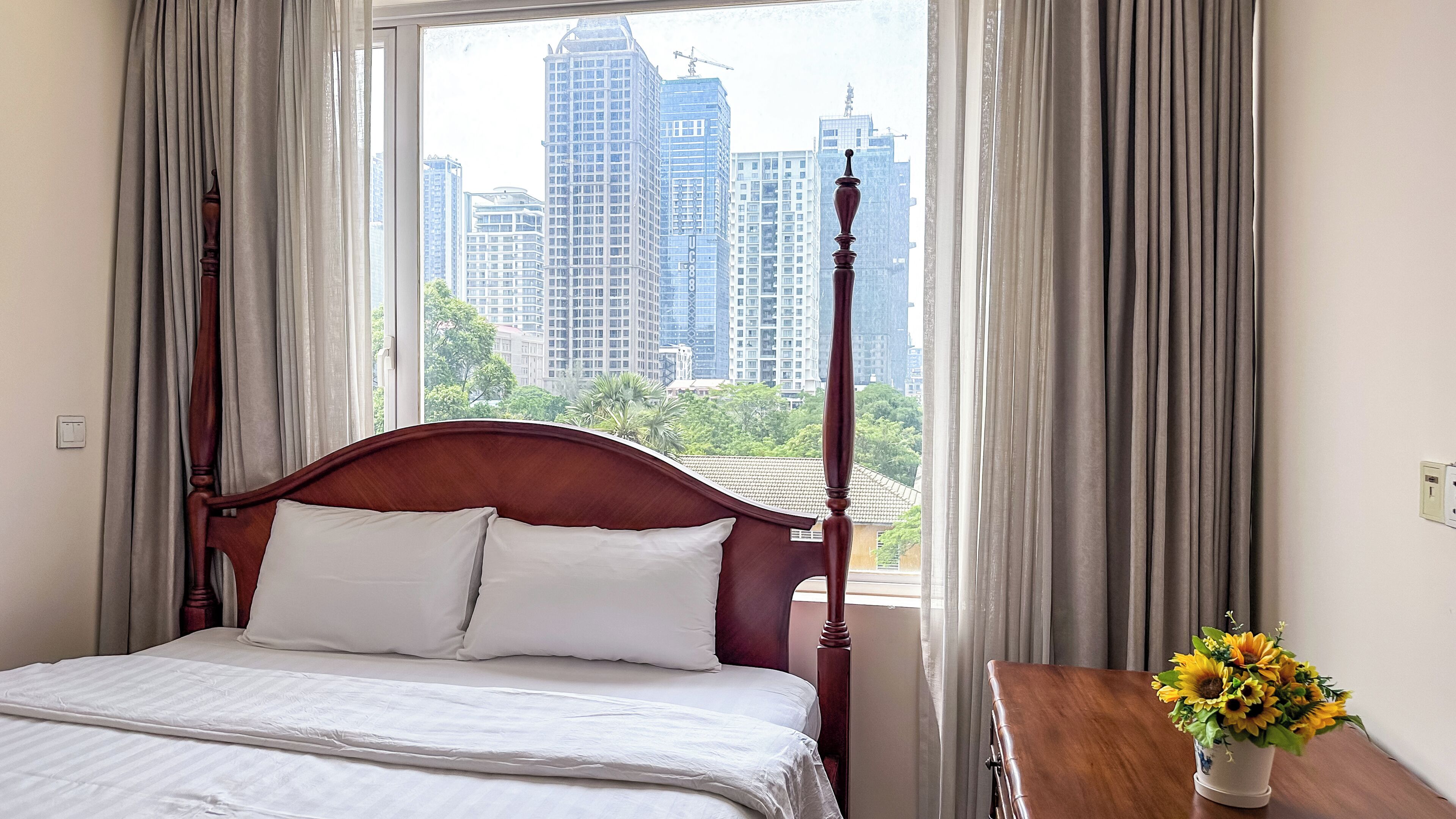 Quarto de hotel com uma janela grande, estrutura de cama de madeira, roupa de cama branca e um vaso com flores sobre uma mesa de cabeceira.