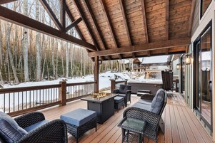 Um deck coberto ao ar livre com móveis de vime, uma lareira e vista para uma floresta nevada.