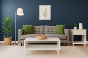 Sala de estar com sofá cinza, almofadas verdes, mesa de centro branca, abajur, planta emoldurada e mesa lateral com um vaso.