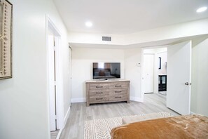 Um quarto com uma cama, uma cômoda, uma televisão e uma porta que dá para outro espaço.