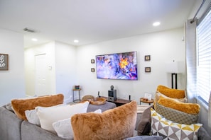 Uma sala de estar moderna com uma televisão de tela plana, um sofá com almofadas estampadas, uma mesinha lateral e uma peça decorativa na parede.