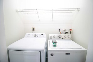 Uma lavanderia com duas máquinas de lavar brancas, uma Samsung e uma Maytag, e um varal em cima.