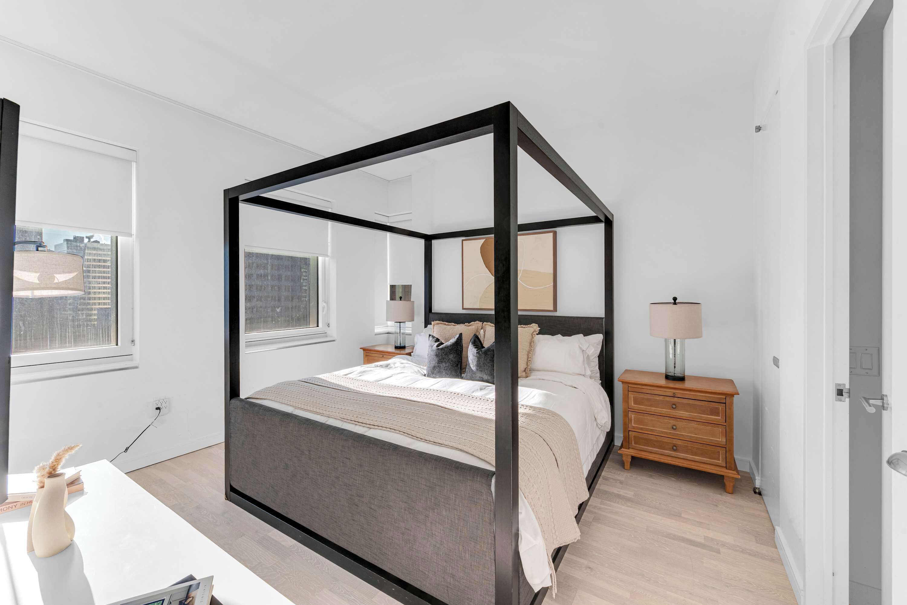 Quarto moderno com cama de dossel, mesa de cabeceira, luminária e vista da cidade através de grandes janelas.
