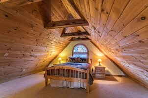 Um quarto de sótão aconchegante com teto de madeira inclinado, uma cama com cabeceira, duas luminárias de cabeceira e uma janela com vitrais.
