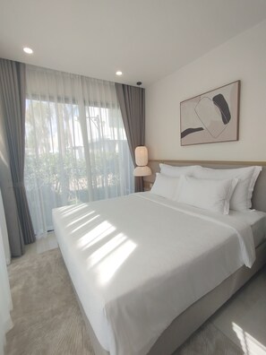 Quarto de hotel com cama grande, luminária de cabeceira e vista para o verde pela janela.