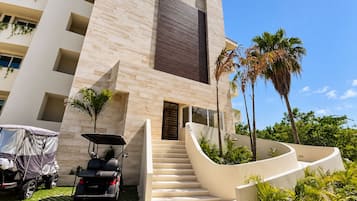 Edifício moderno com escada que leva à entrada, cercado por palmeiras e uma área coberta para veículos.