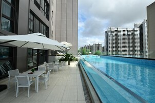 Área de piscina no terraço, com guarda-sóis brancos, espreguiçadeiras e vista para edifícios altos.