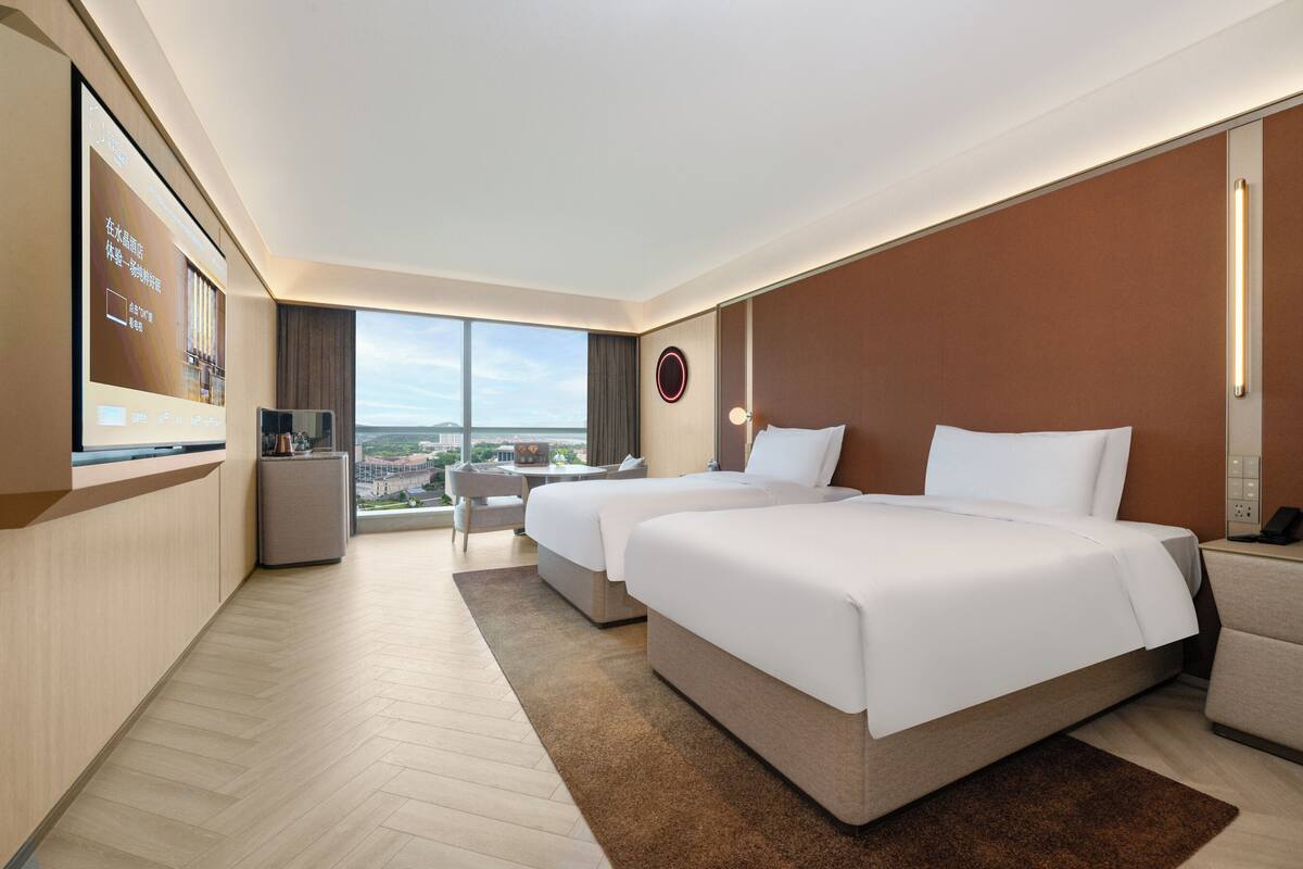 Quarto de hotel com duas camas, uma escrivaninha, televisão e uma janela grande com vista para a cidade.