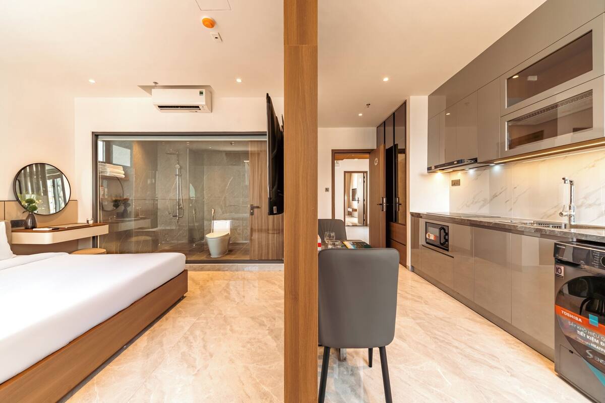 Quarto de hotel moderno com cama, área de estar, chuveiro, cozinha equipada com micro-ondas e uma janela grande.