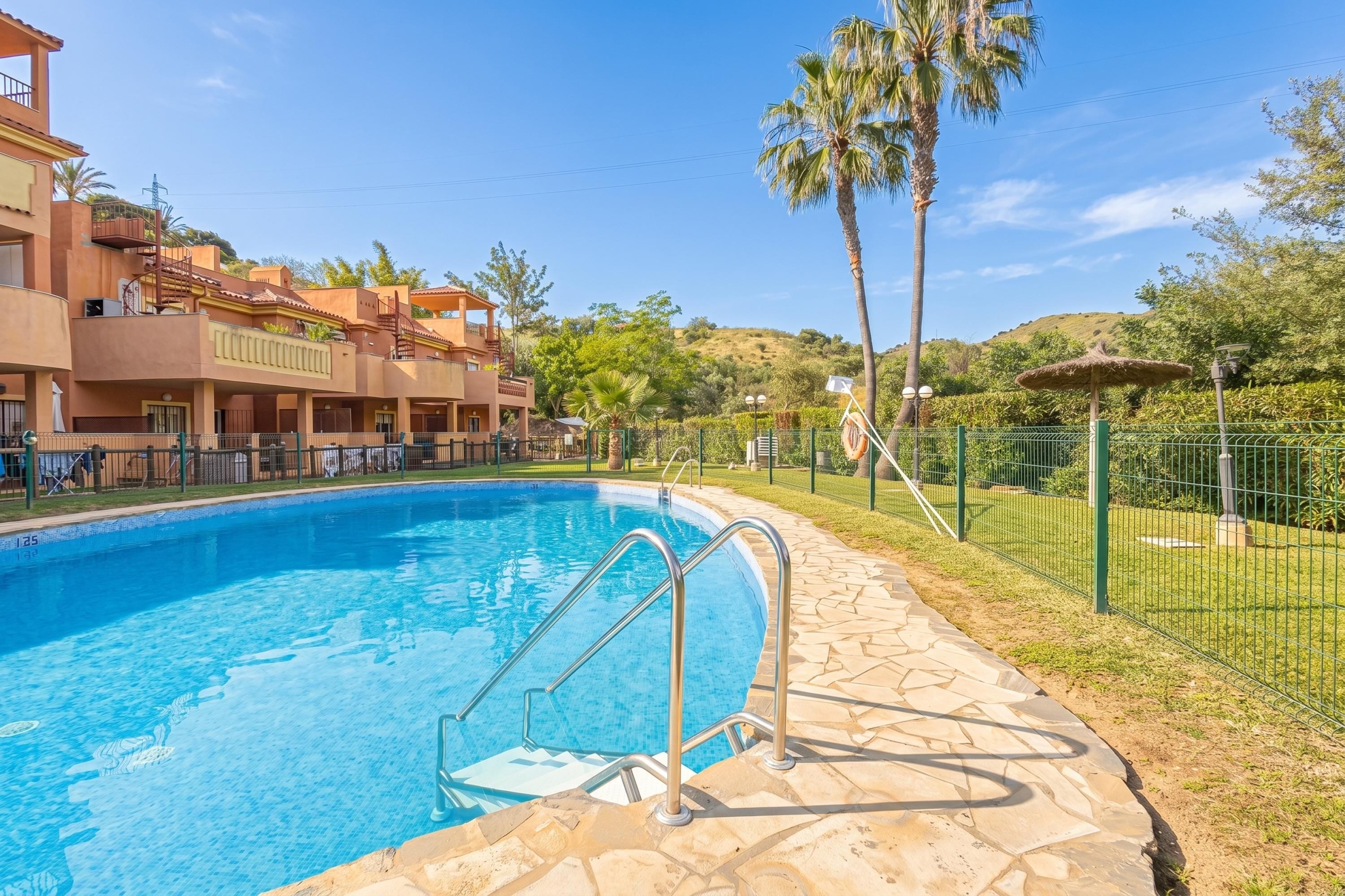 Piscina com deck de pedra, cercada por uma grade e palmeiras, com um edifício ao fundo.
