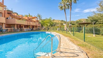 Piscina com deck de pedra, cercada por uma grade e palmeiras, com um edifício ao fundo.