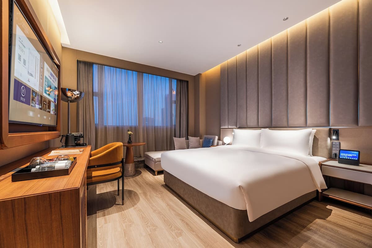 Quarto de hotel moderno com uma cama grande, uma escrivaninha, uma cadeira, uma máquina de café e um painel de controle digital.