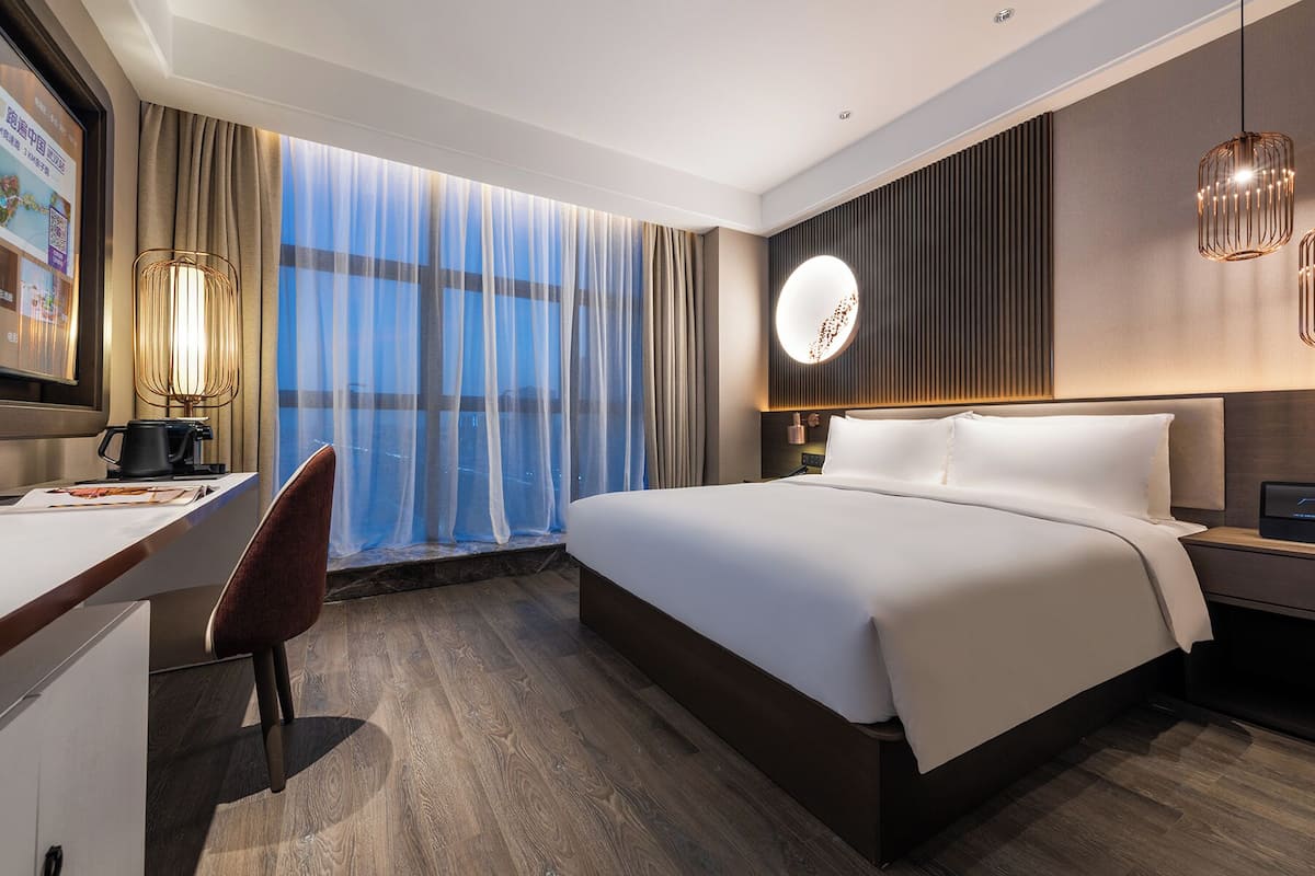 Quarto de hotel moderno com uma cama grande, uma escrivaninha com cadeira, uma luminária redonda na parede e uma janela ampla com cortinas.