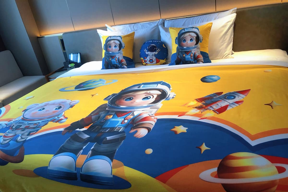 Uma cama com edredom decorado com tema espacial, almofadas e uma mesa de cabeceira com abajur.