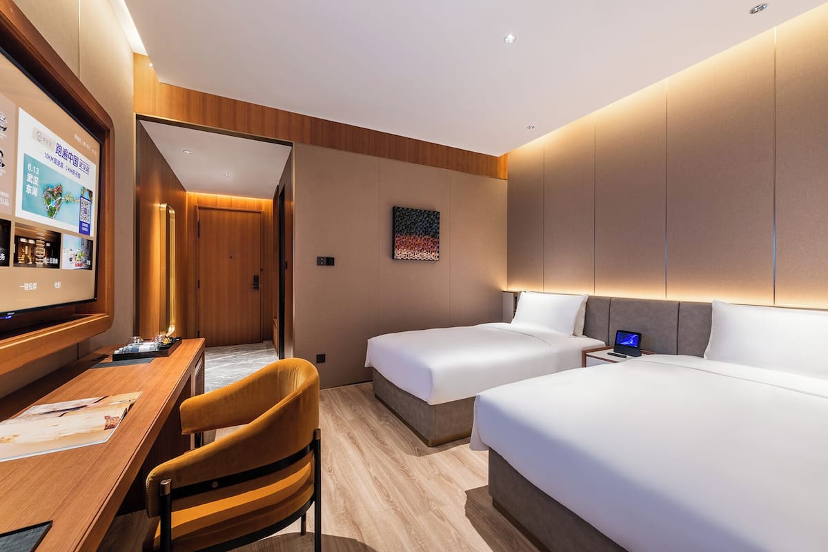 Quarto de hotel com uma escrivaninha, duas camas, uma cadeira, uma televisão e uma porta que dá para outro cômodo.