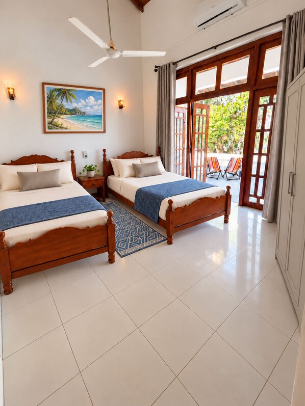 Quarto de hotel com duas camas de madeira, um ventilador de teto, um quadro com tema praiano e vista para o exterior por uma porta de vidro.