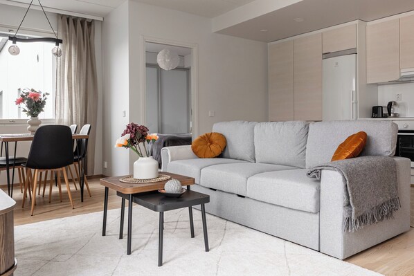 Uma sala de estar moderna com um sofá cinza, uma mesinha de centro de madeira e um espaço de refeições com um vaso branco e flores.