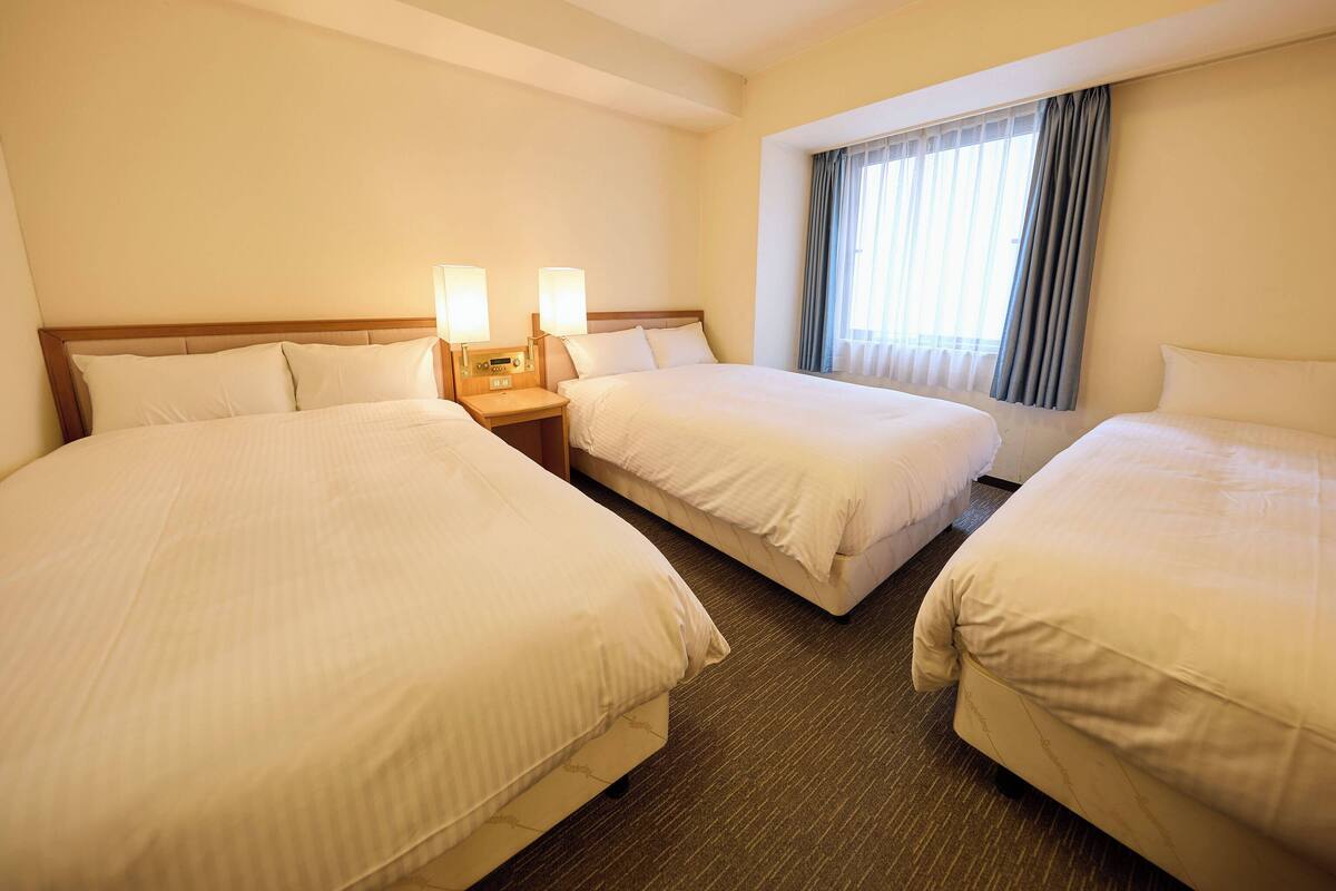 Quarto de hotel com três camas, uma mesa de cabeceira com abajures, uma janela com cortinas e piso coberto por carpete.