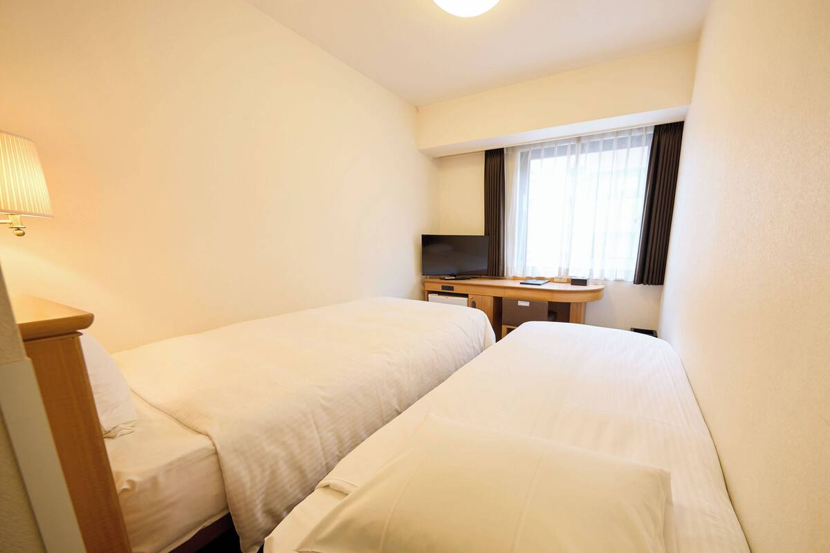 Quarto de hotel com duas camas, uma escrivaninha, televisão e janela com cortinas.