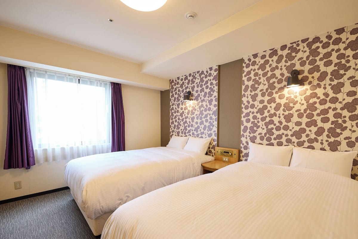 Quarto de hotel com duas camas, uma janela com cortinas, uma luminária fixada na parede e um termostato.