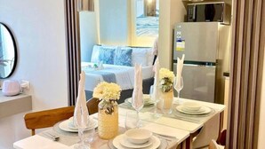 Uma sala de jantar com uma mesa posta para quatro pessoas, uma cozinha ao fundo e uma cama visível pela janela.