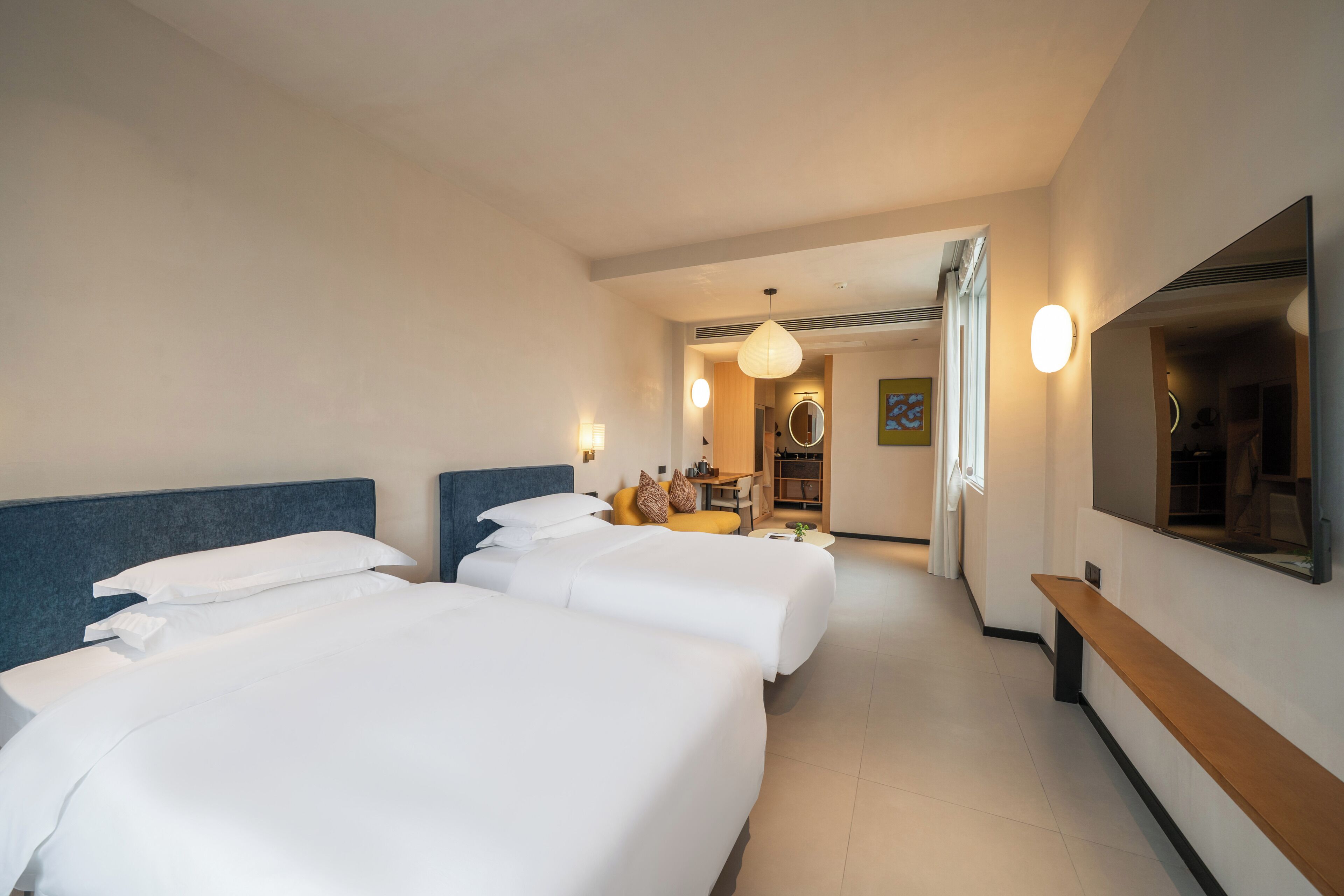 Quarto de hotel com duas camas, um banco, um espelho e uma área de refeições ao fundo.
