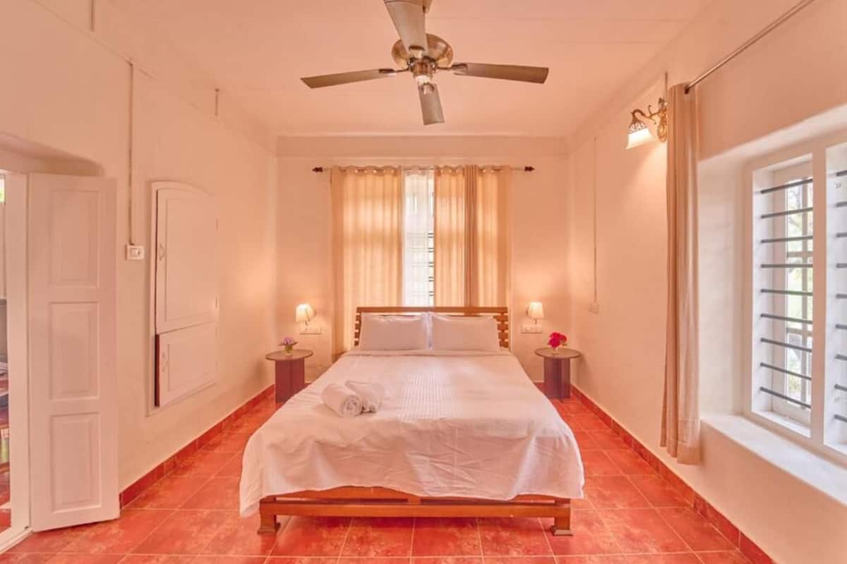 Quarto de hotel com cama, ventoinha no teto, janela com cortinas e mesa de cabeceira com luminária.