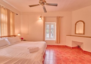 Quarto de hotel com cama, ventoinha no teto, janela com cortinas, espelho e chão ladrilhado.