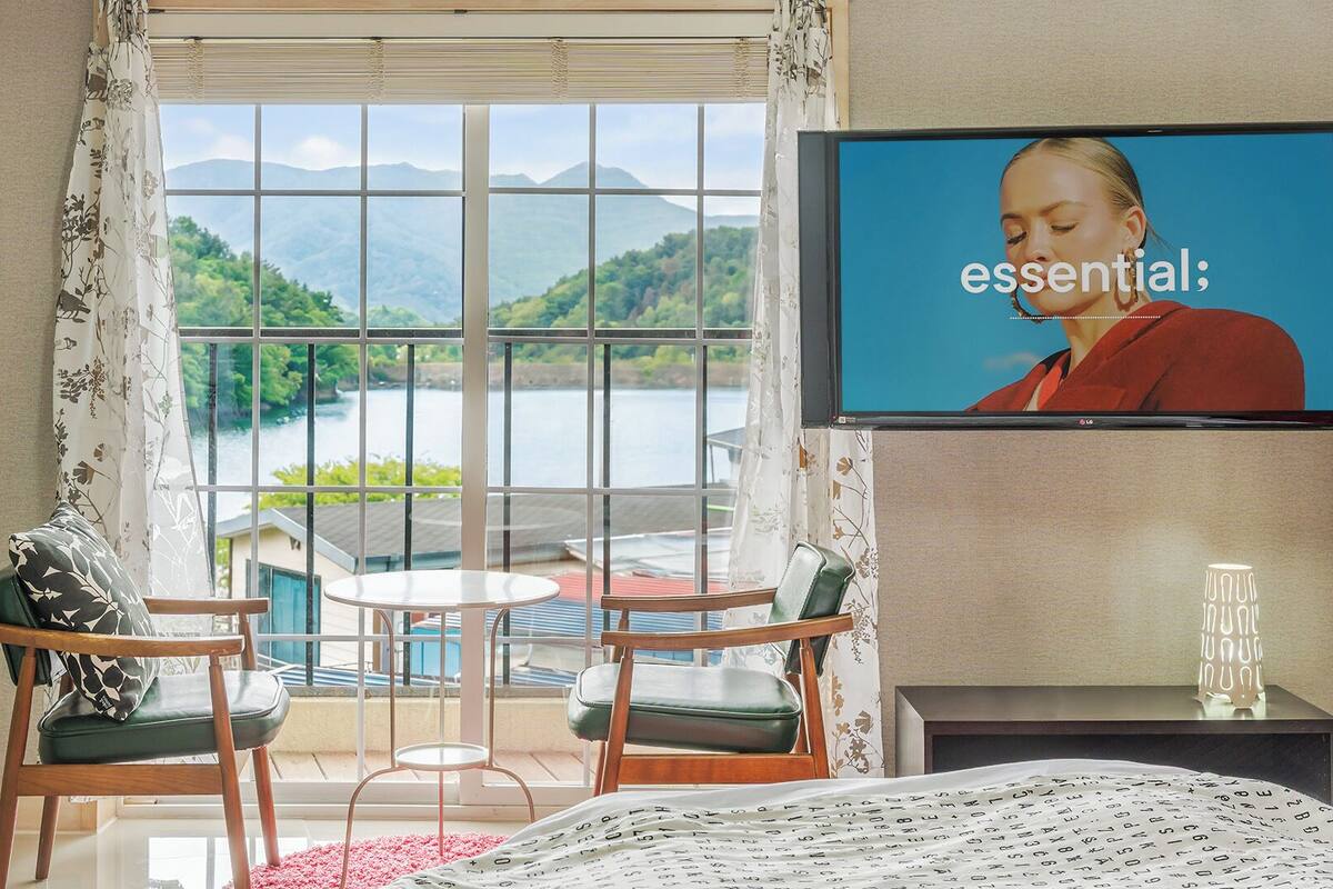 Quarto de hotel com uma janela grande, uma cama, uma TV e uma pequena mesa com cadeiras.