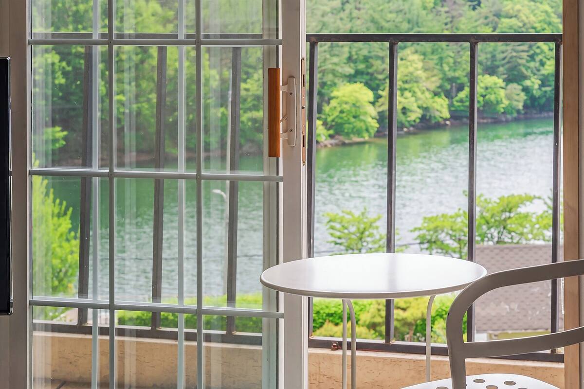 Quarto com mesa, cadeira e janela que dá para um rio e vegetação.