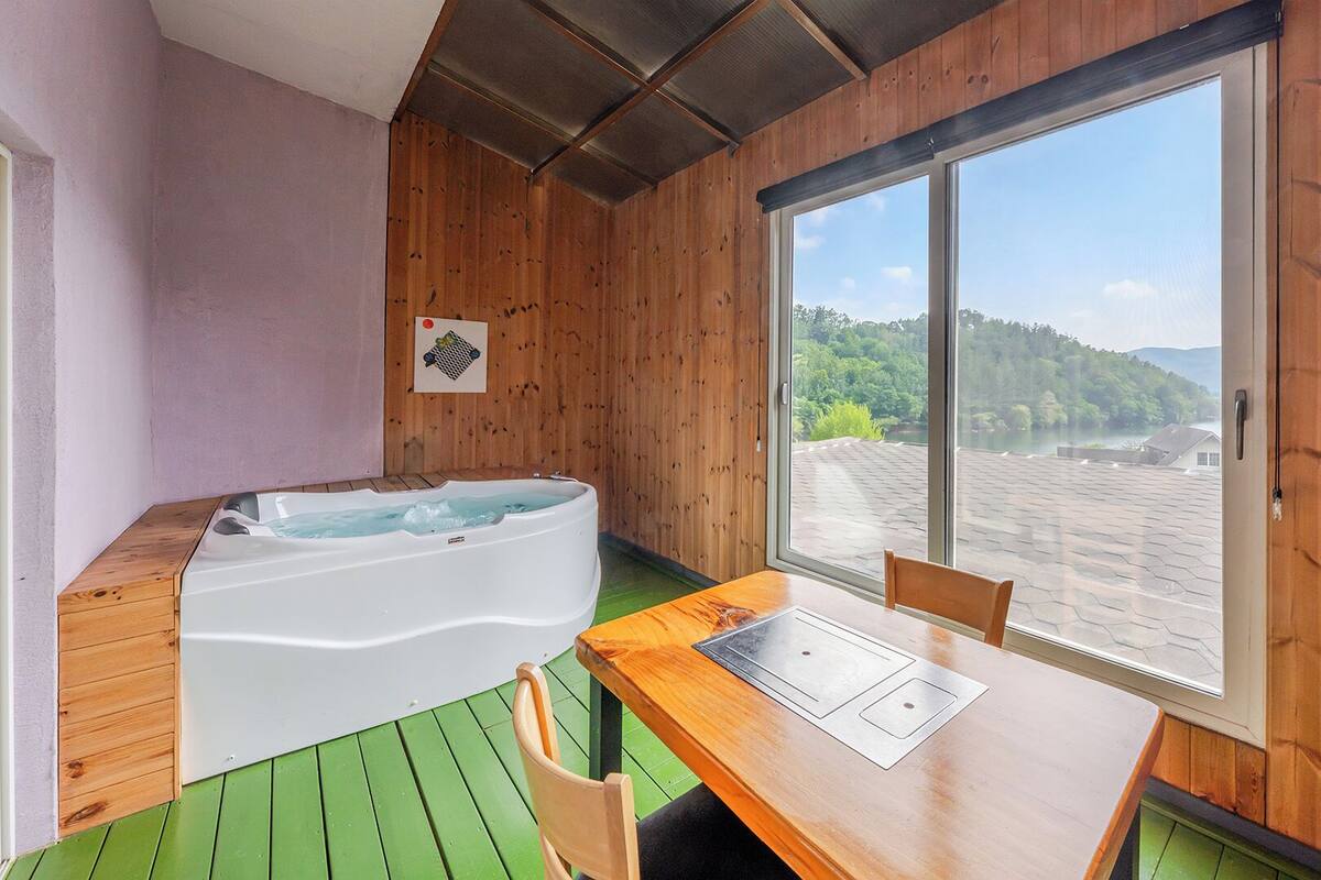 Quarto com decoração em madeira, banheira de hidromassagem, mesa com cadeiras e uma grande janela com vista para uma área arborizada.