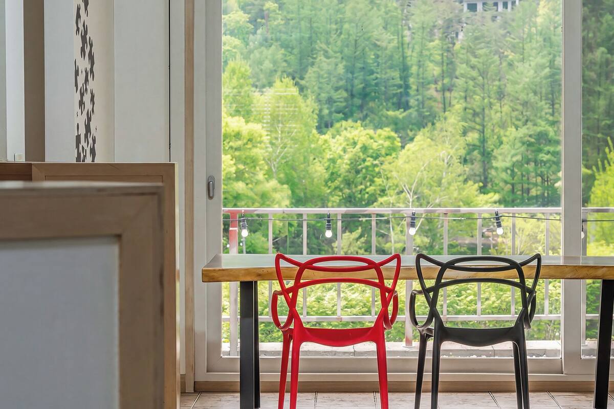 Uma varanda com mesa de madeira e duas cadeiras, uma vermelha e outra preta, com vista para uma área arborizada.