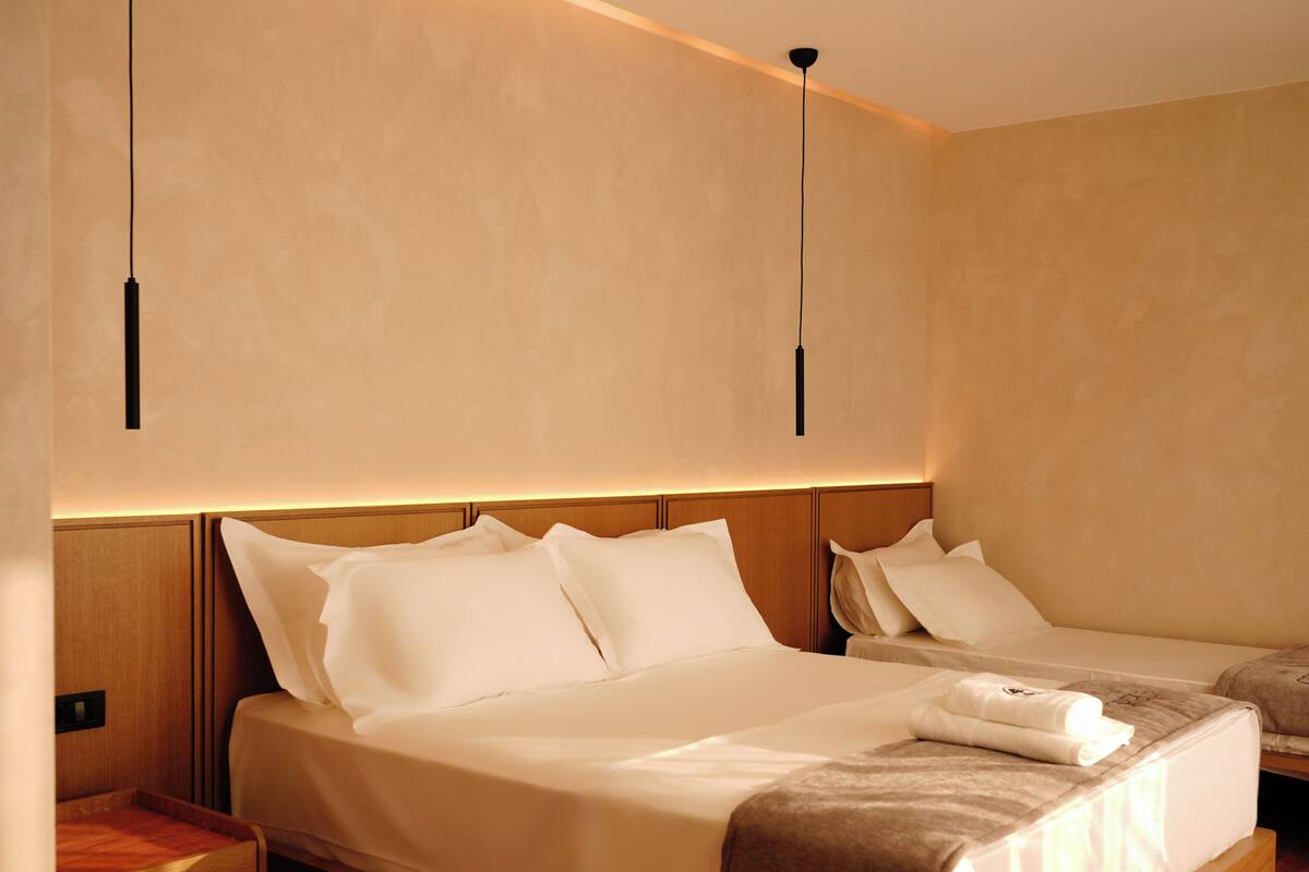 Quarto de hotel com uma cama grande, uma cama menor, cabeceira de madeira e iluminação pendente.