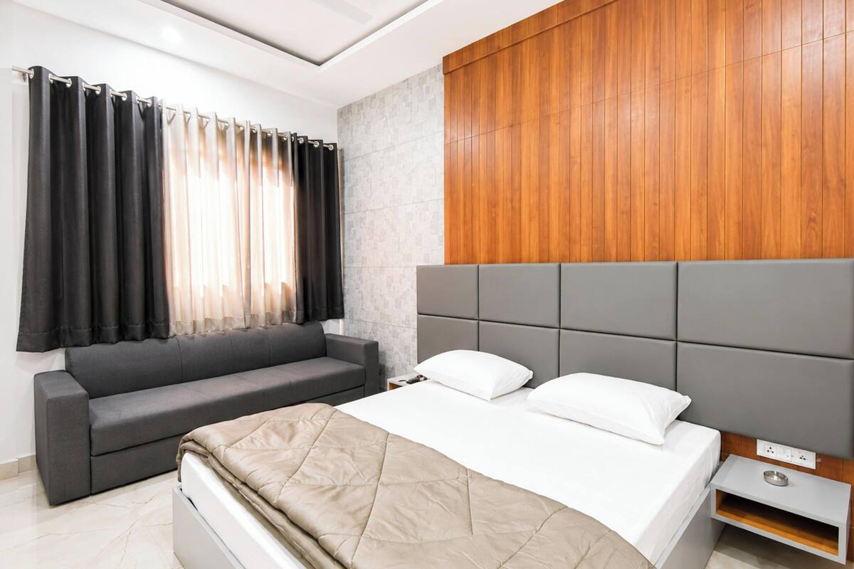 Quarto moderno com cama, sofá cinzento, painel de parede em madeira e janela com cortinas.