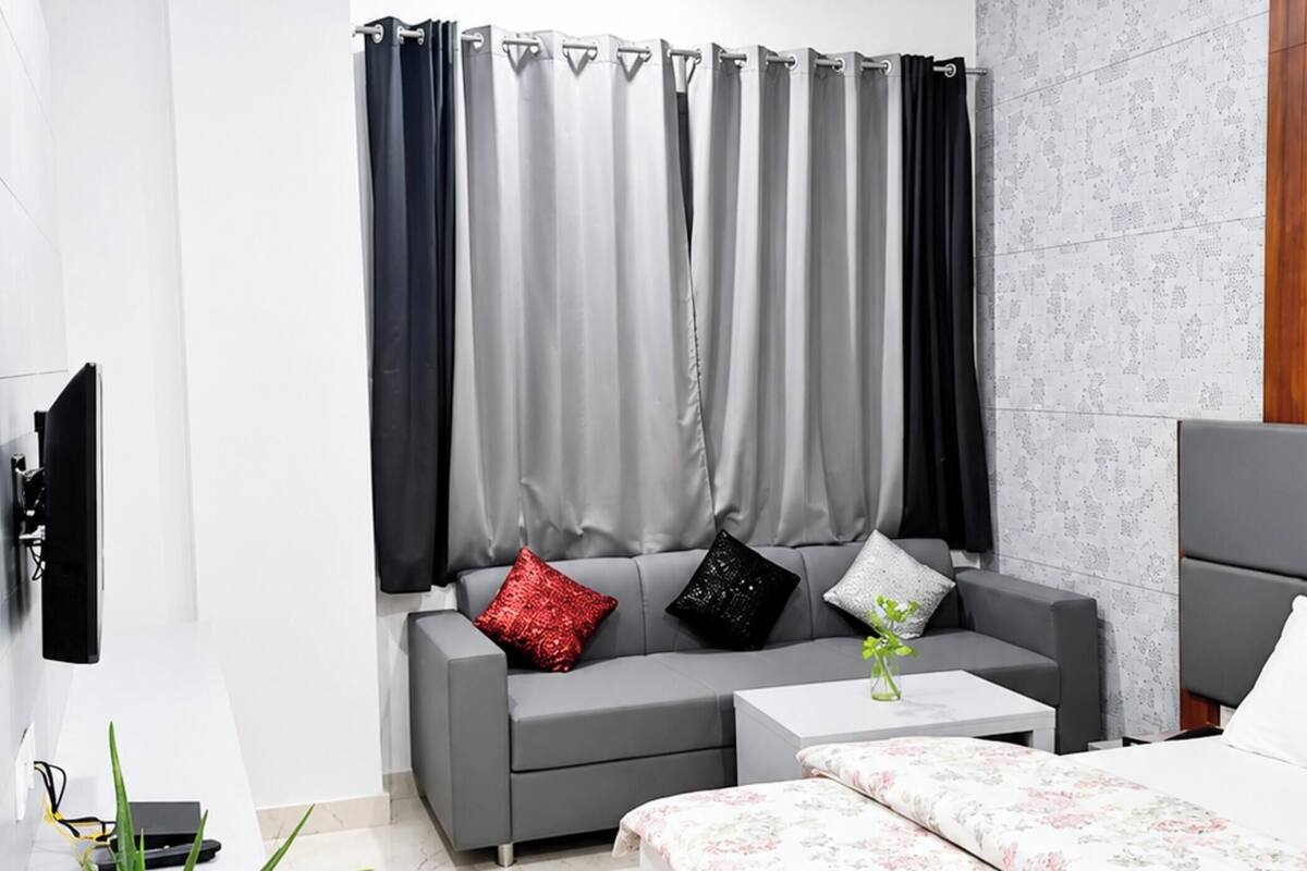Um quarto moderno com sofá cinza, cama com roupa de cama florida, mesa de cabeceira branca, TV fixada na parede e ventoinha de teto.