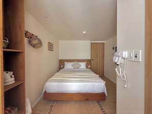 Quarto com estrutura de cama de madeira, roupa de cama branca, secador de cabelo fixado na parede e prateleira com toalhas dobradas.