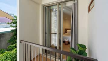 Varanda com grade de metal, porta de vidro deslizante que dá para um quarto com cama e mesa de cabeceira, e uma pequena planta à direita.