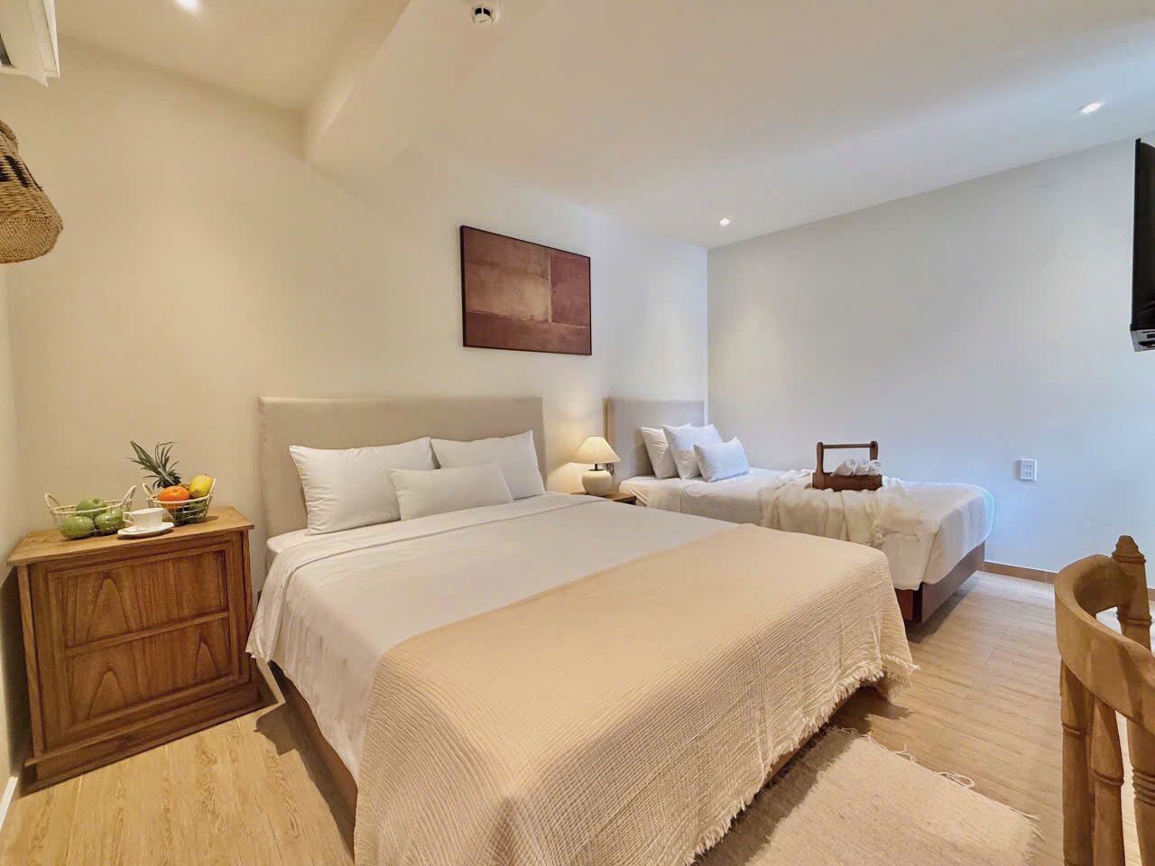 Quarto com uma cama grande, uma cama menor, uma mesinha de cabeceira de madeira, um abajur e uma cesta de frutas.