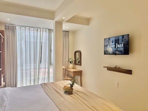 Quarto de hotel com cama, escrivaninha com espelho, televisão na parede e janela com cortinas.