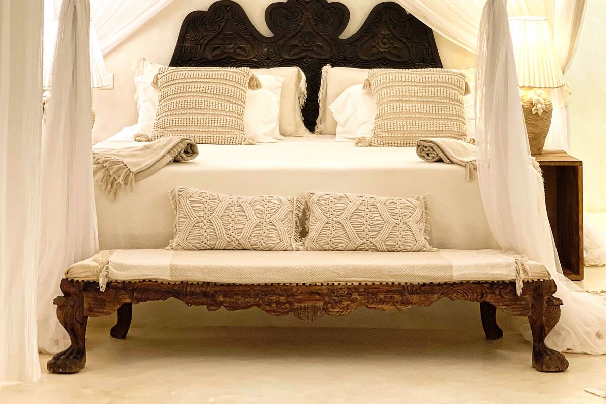 Uma cama com dossel, cabeceira de madeira e almofadas estampadas.