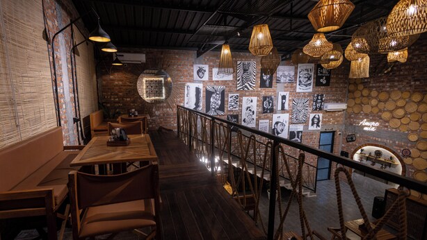 Um restaurante com mesas e cadeiras de madeira, uma parede de tijolos com obras de arte e luminárias de vime penduradas.