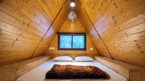 Quarto com teto inclinado, cama com roupa de cama branca, duas almofadas e um cobertor marrom. Há uma janela com vista para árvores.