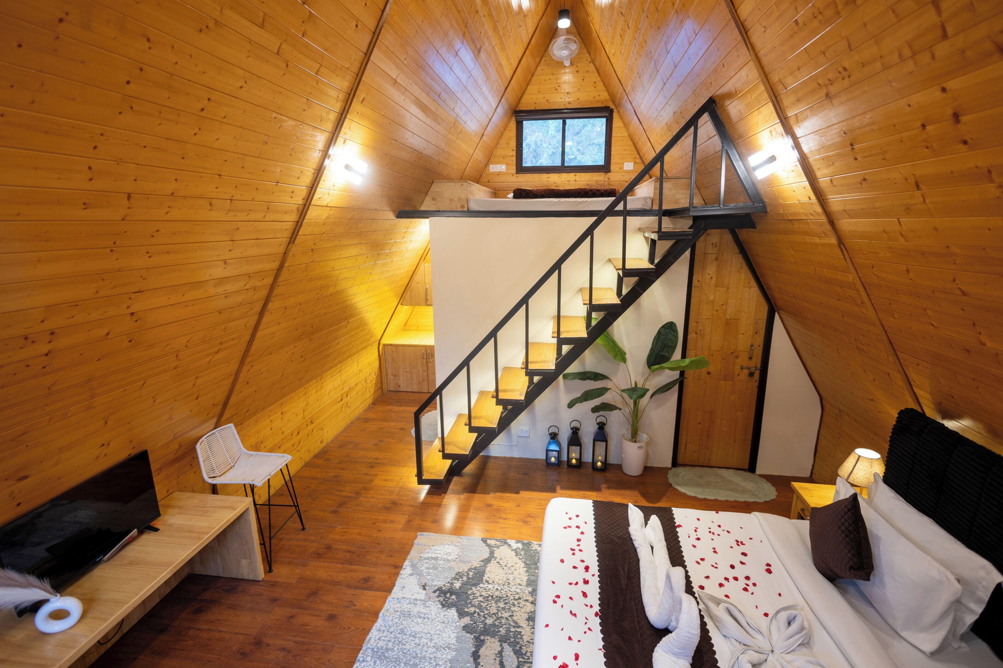 Um quarto de sótão acolhedor com teto inclinado de madeira, uma escada, uma cama com cabeceira decorativa, uma escrivaninha com cadeira e uma pequena janela.