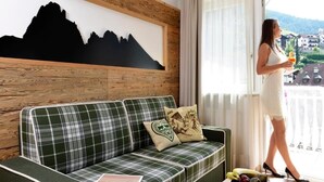 Quarto com sofá xadrez, arte com silhueta de montanhas, janela com vista para casas e mesa com frutas.