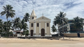Uma igreja com um campanário, rodeada por palmeiras e uma praia de areia.