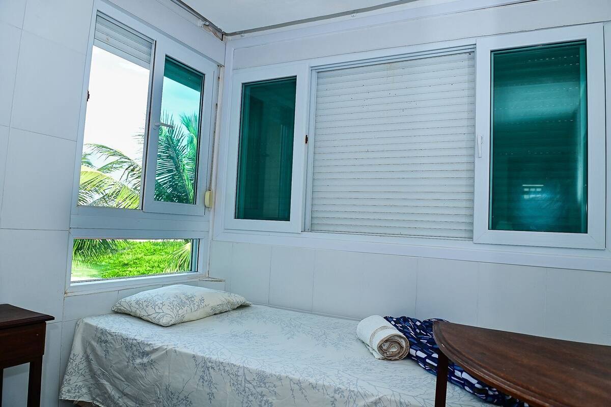 Um quarto pequeno com cama, duas janelas com persianas e vista para o verde lá fora.