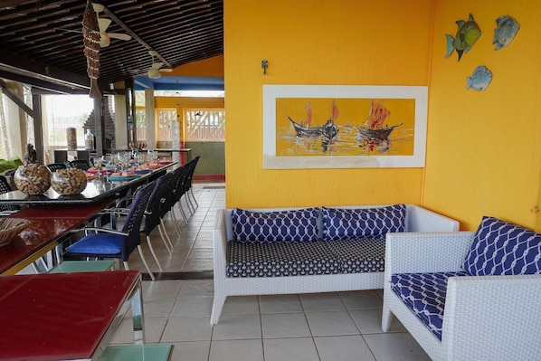 Restaurante com parede amarela, pintura de barcos, móveis de vime e chão de azulejos.