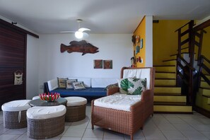 Uma sala de estar com sofá, dois pufes redondos de vime, uma cadeira de vime, ventilador de teto, decoração de peixe e uma escada.