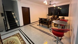Uma sala de estar com escada, mesa de jantar com cadeiras, um banquinho vermelho e um ventilador de teto.